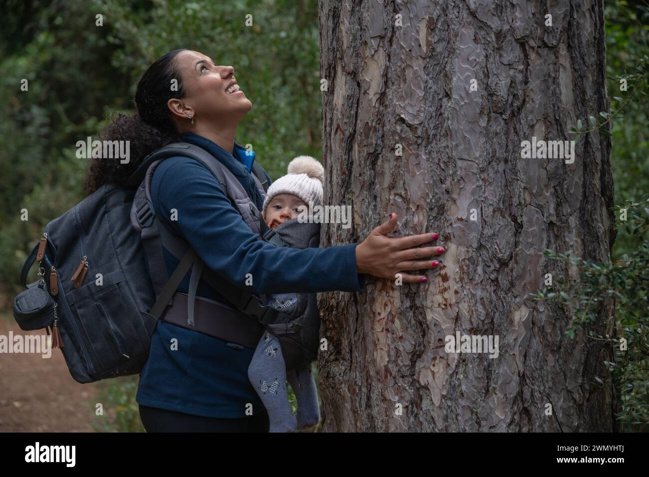 A smiling mother wearing a backpack hugs a tree while carrying her baby ...