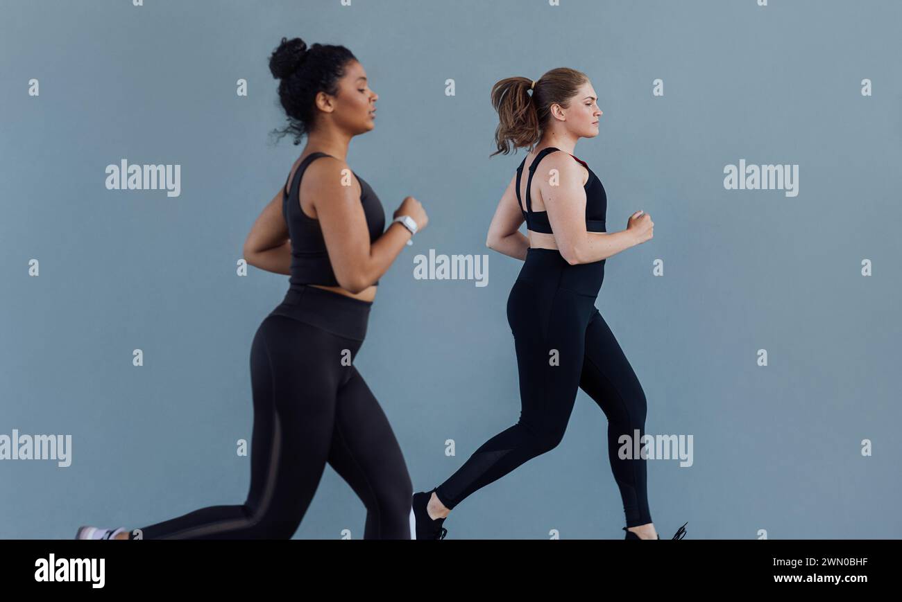 Side view of two plus size females running at grey wall. Two women in ...