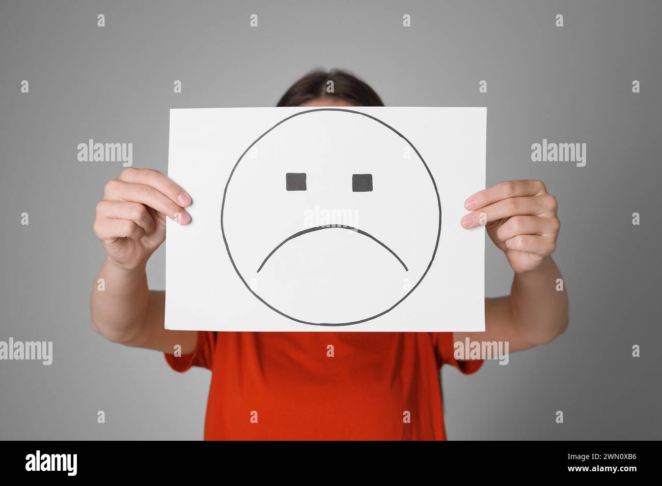 Woman hiding behind sheet of paper with sad face on grey background ...