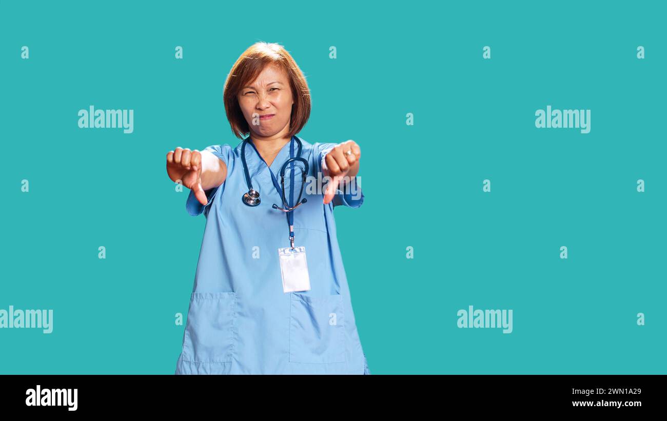 Upset asian nurse showing thumbs down signs while at work, isolated ...
