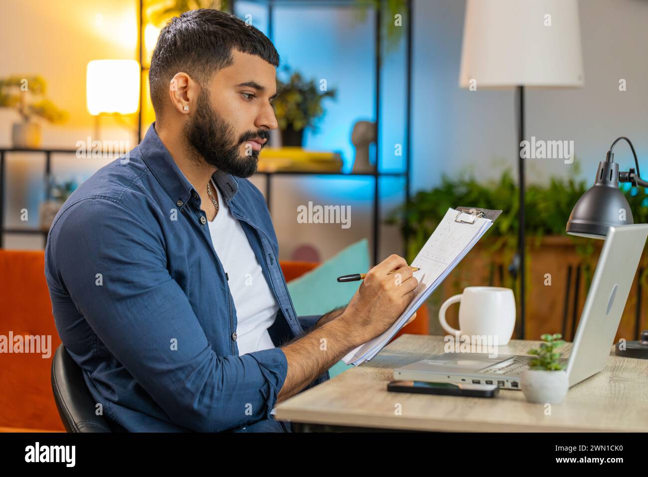 Indian young bearded freelancer making notes on clipboard at home ...
