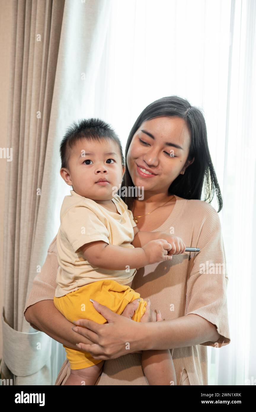 A loving and caring Asian mom is holding her cute little son in the living room. motherhood and ...