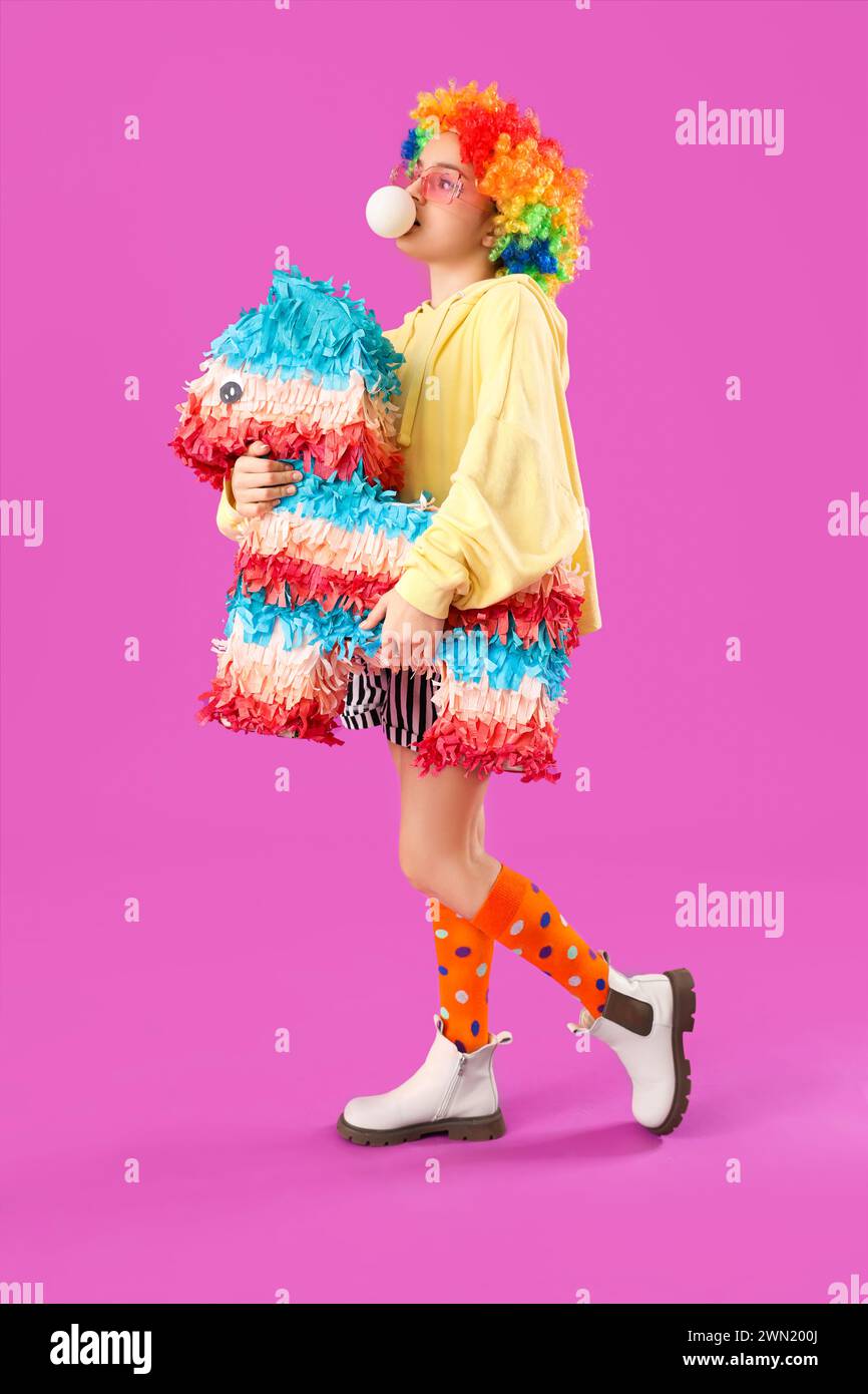 Trendy girl in clown costume with pinata blowing bubble gum on purple ...