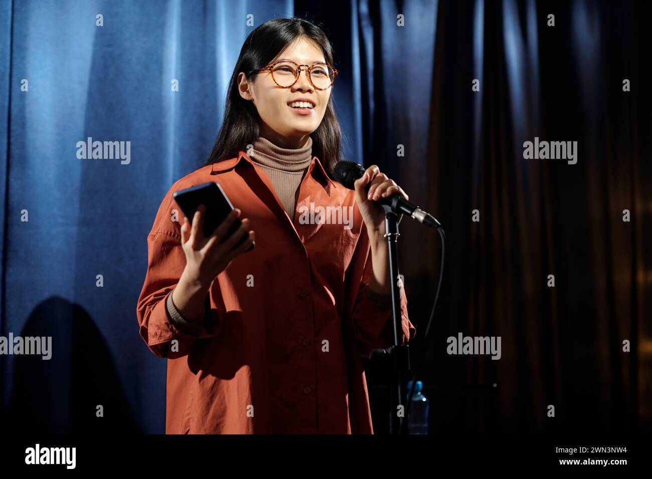 Happy young female comedian with smartphone and microphone standing on ...