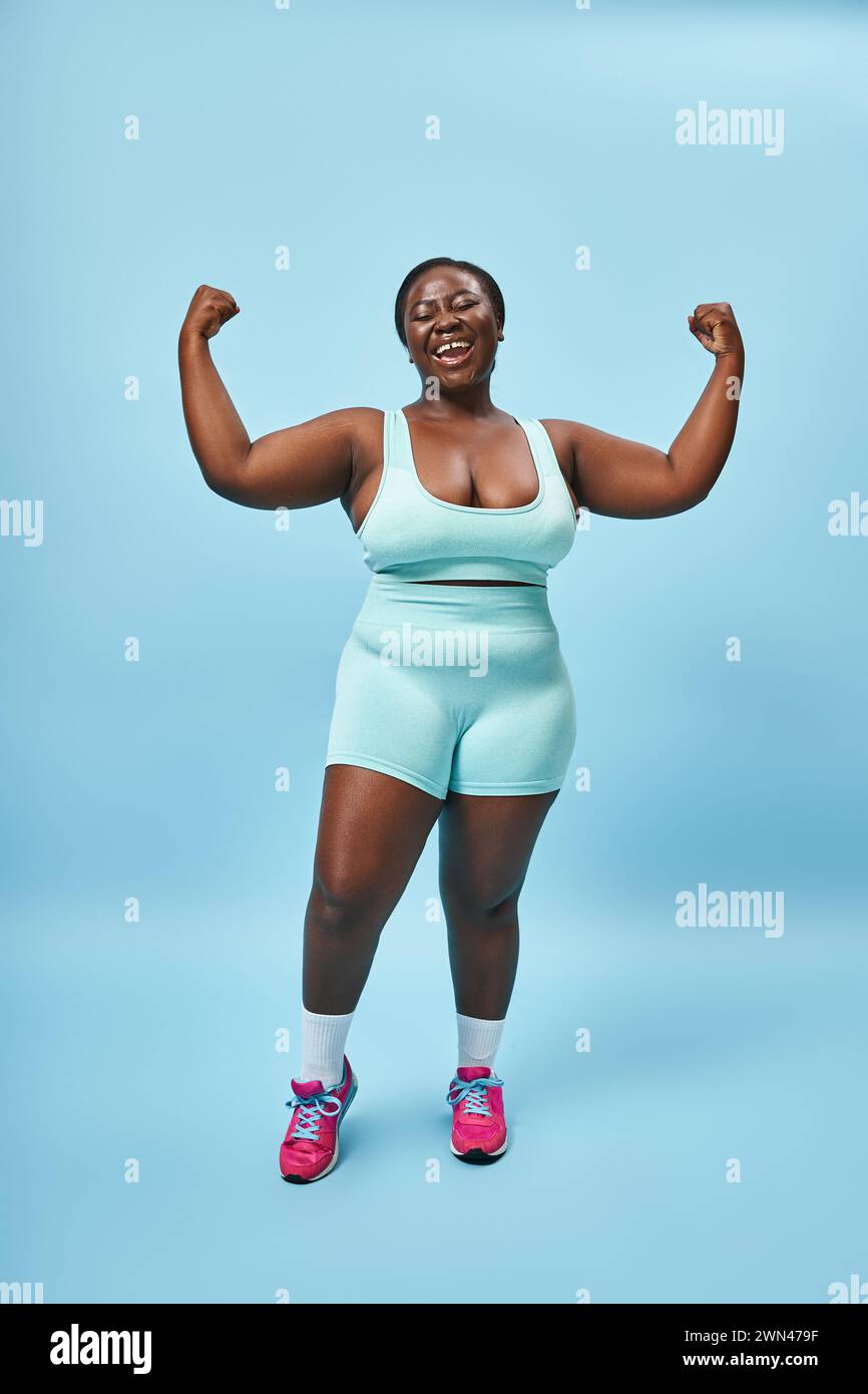 excited plus size woman in active wear flexing her muscles and smiling ...