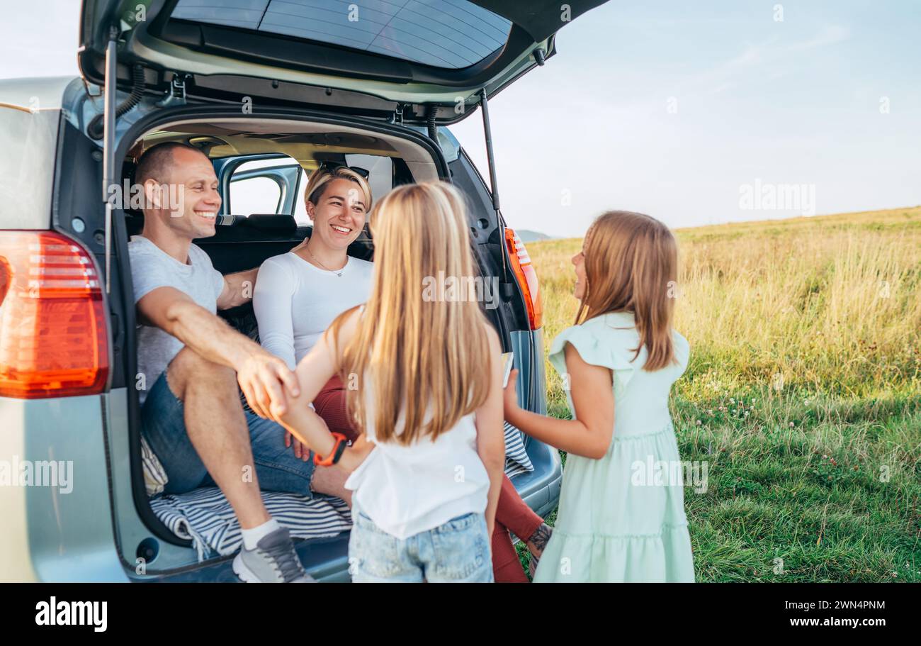 Portrait of Happy young couple with little daughters sitting inside car trunk during auto trop ...