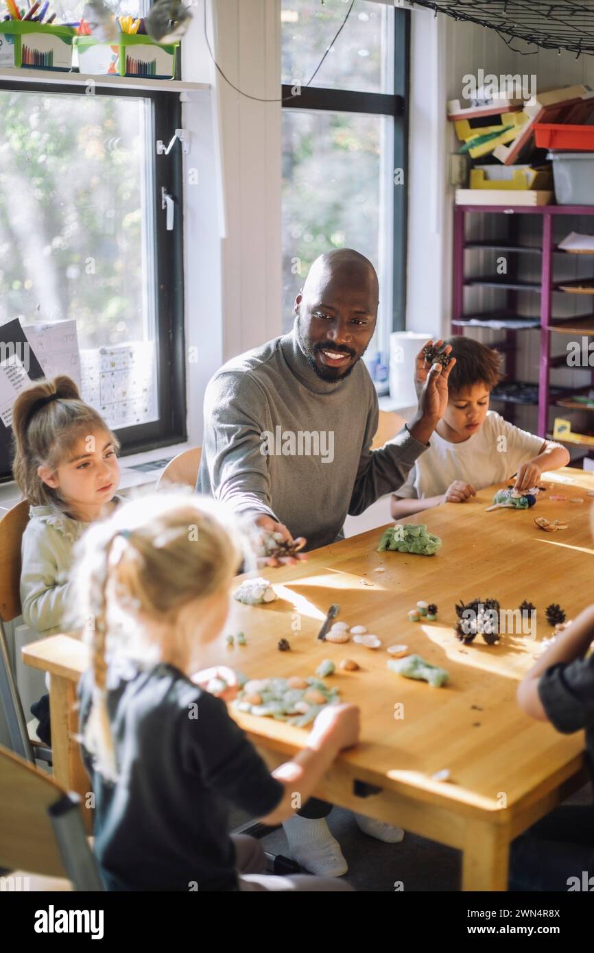 Male teacher playing with kids while sitting at bench during art class ...