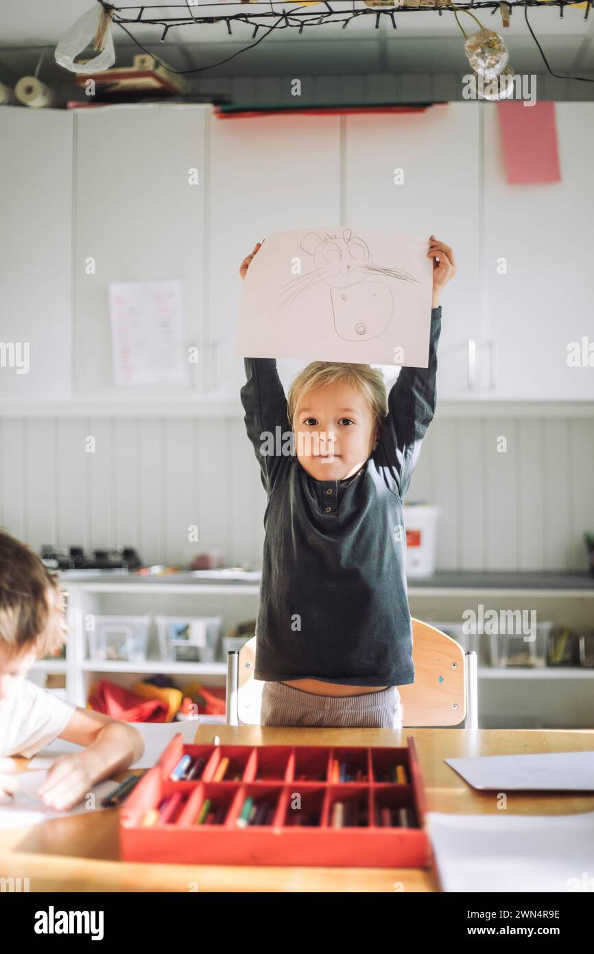 Portrait of girl showing paper with drawing while standing near bench ...