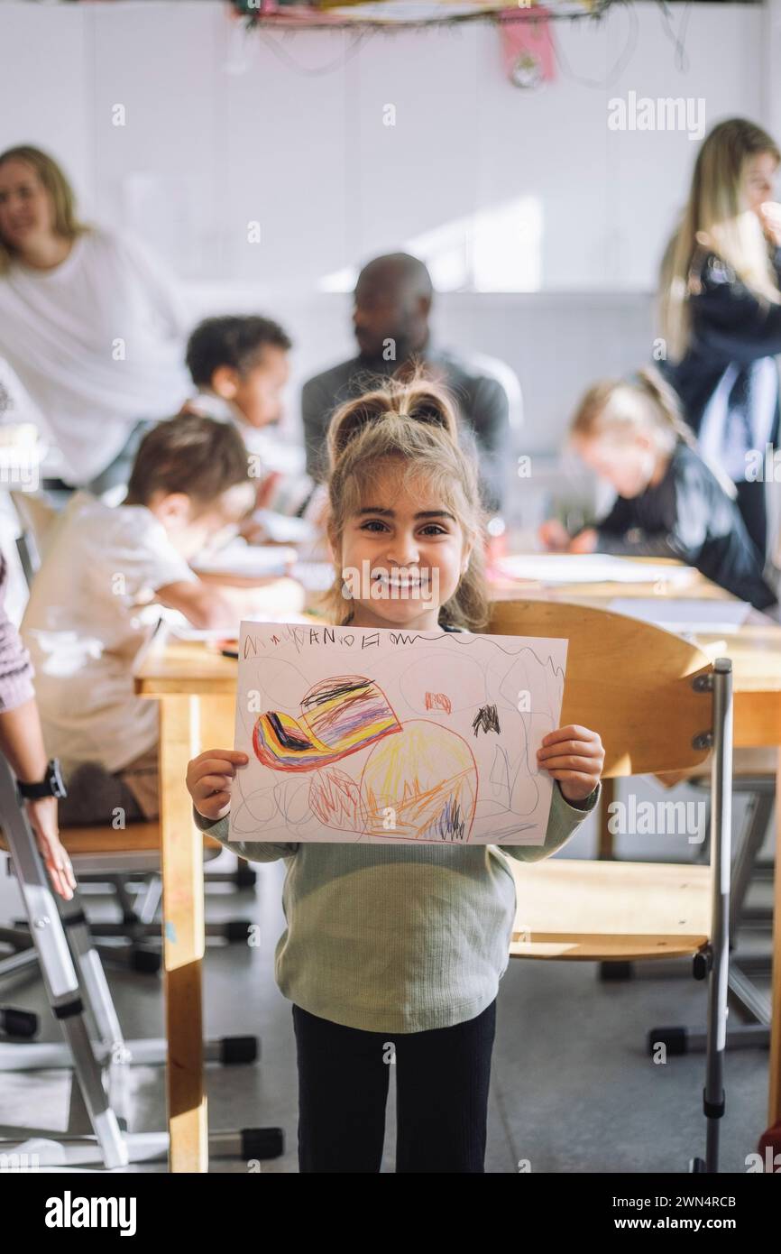 Portrait of smiling girl showing drawing while standing near bench in ...