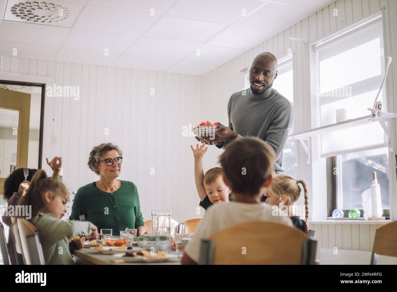 Male teacher serving food to children sitting at dining table during ...