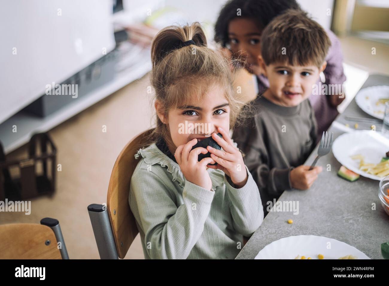 Portrait of girl biting fruit while sitting at dining table with mates ...