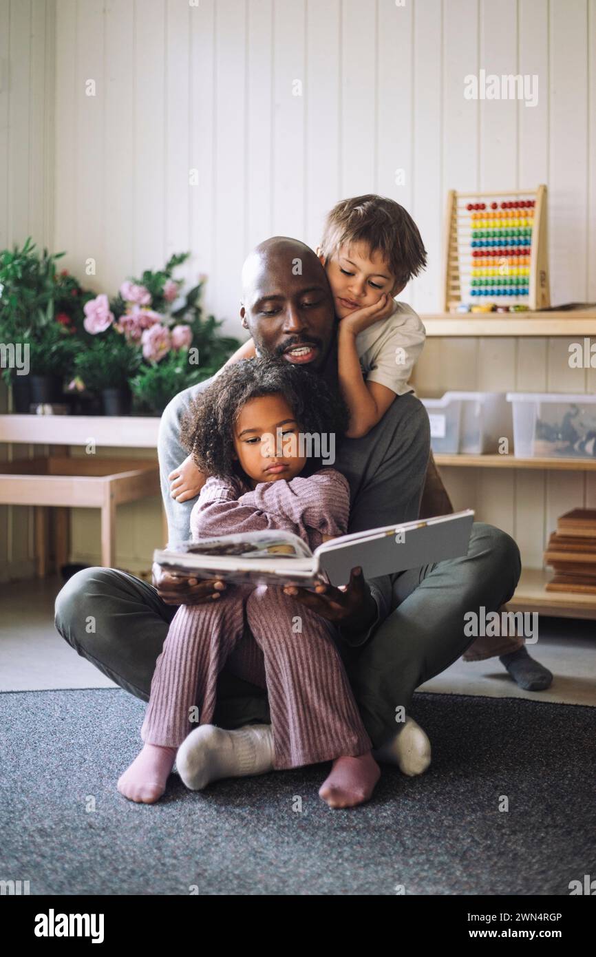 Male teacher reading book to preschool kids while sitting cross-legged ...