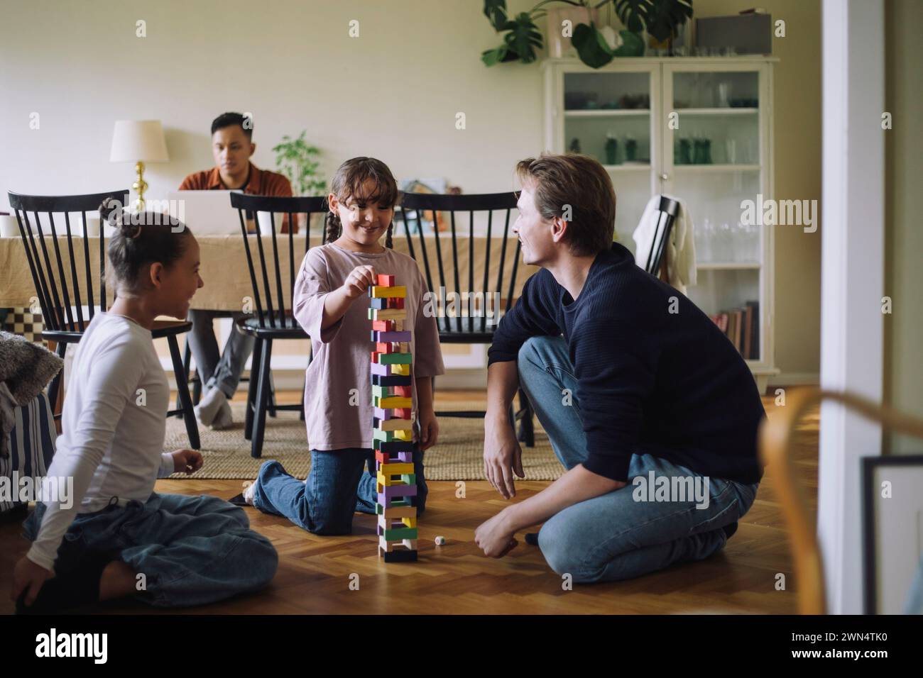 Smiling girl stacking Jenga blocks while kneeling on floor by father ...
