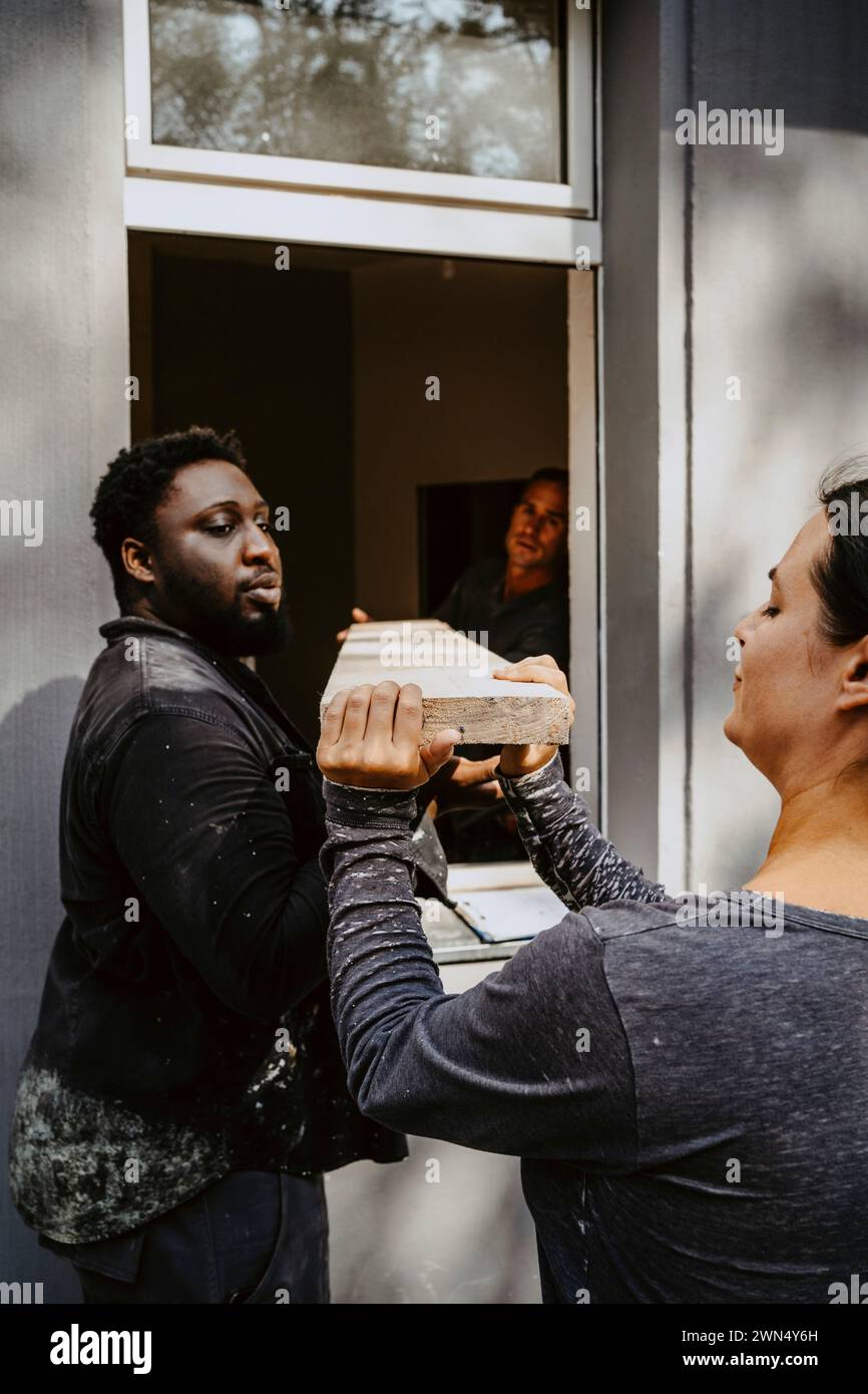 Male and female construction workers passing plank through window of ...