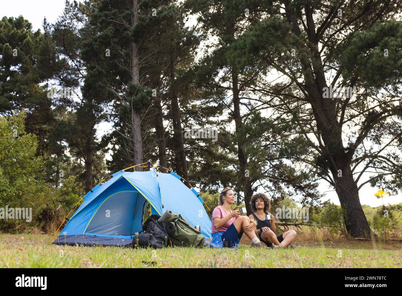 Two women relax beside a blue tent in a grassy outdoor setting with ...