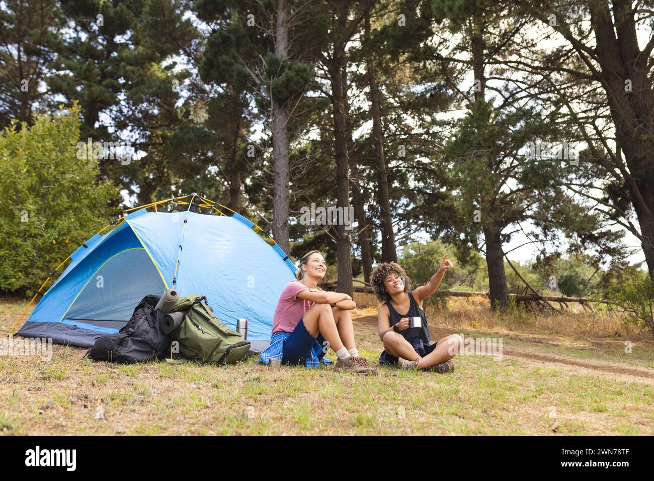 Two women relax beside a blue tent in a wooded campsite with copy space ...