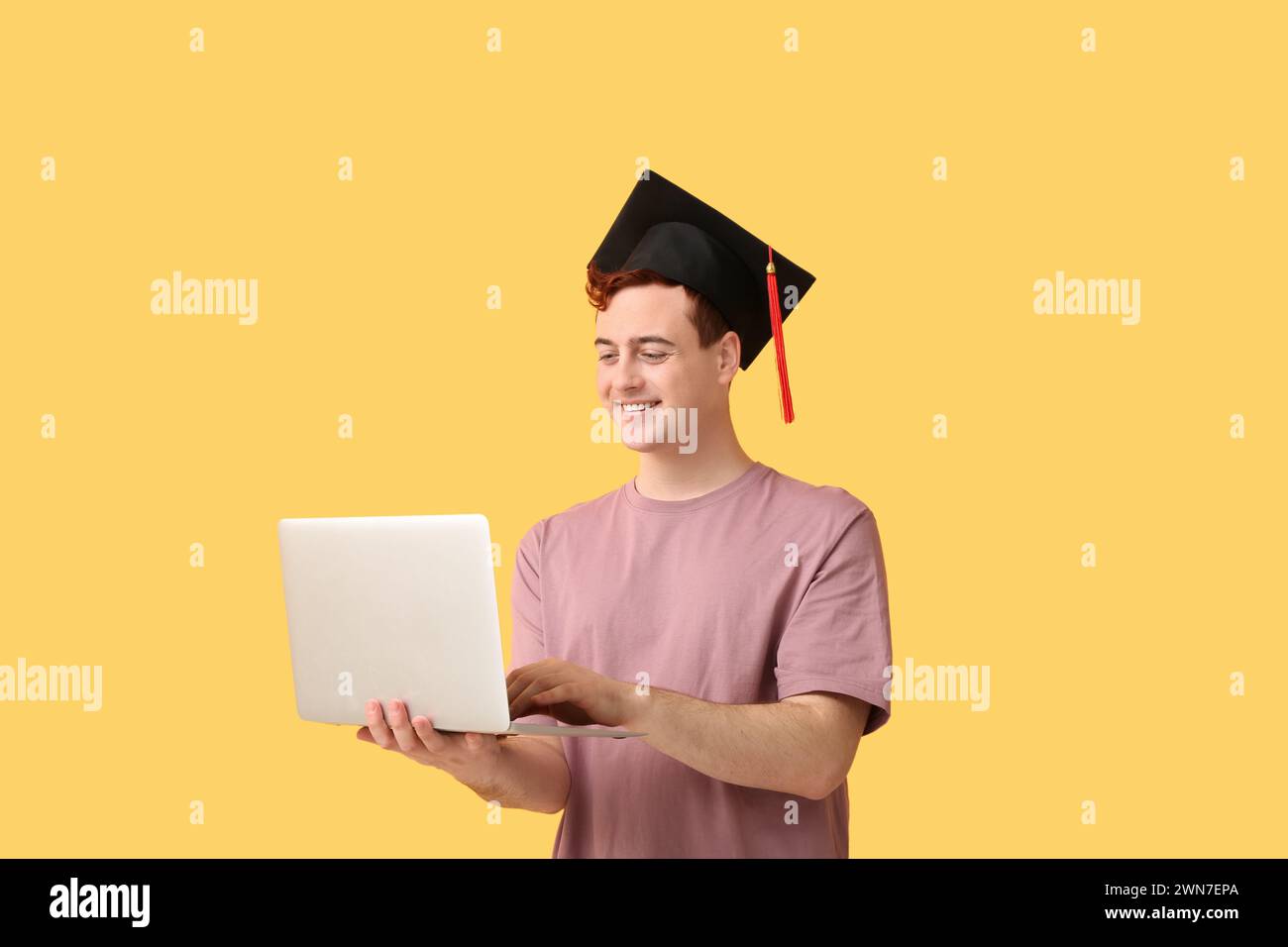 Male student in mortar board with modern laptop on yellow background ...