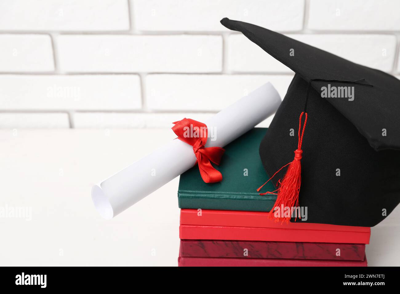 Mortar board with diploma and books on table near white brick wall ...