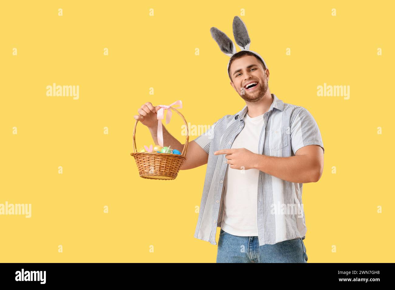 Handsome man in bunny ears pointing at Easter eggs on yellow background ...