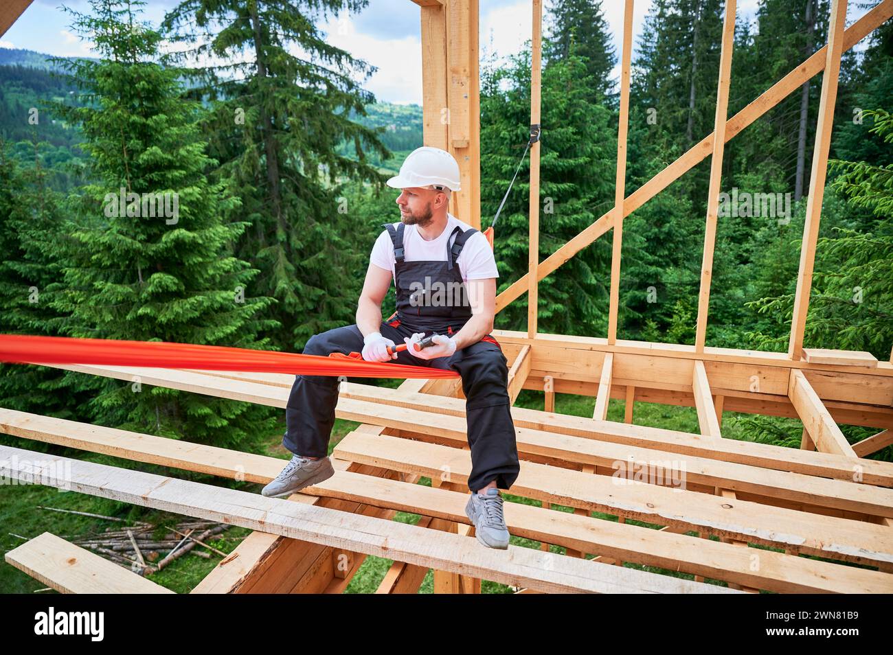 On construction site of wooden-framed house, carpenter takes break in hammock, donning ...