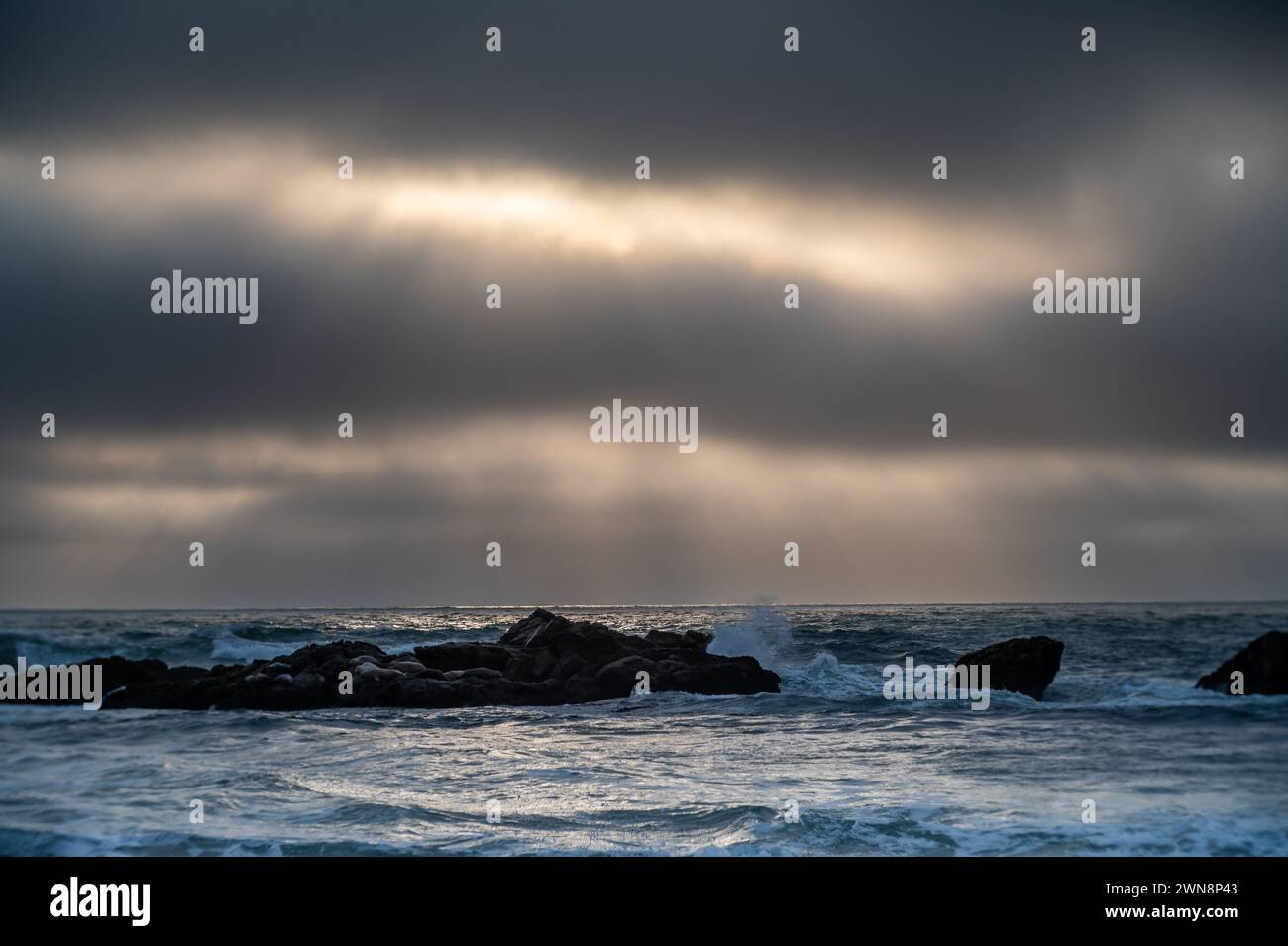 Sun rays coming through gray clouds over ocean waves and coastal rocks ...