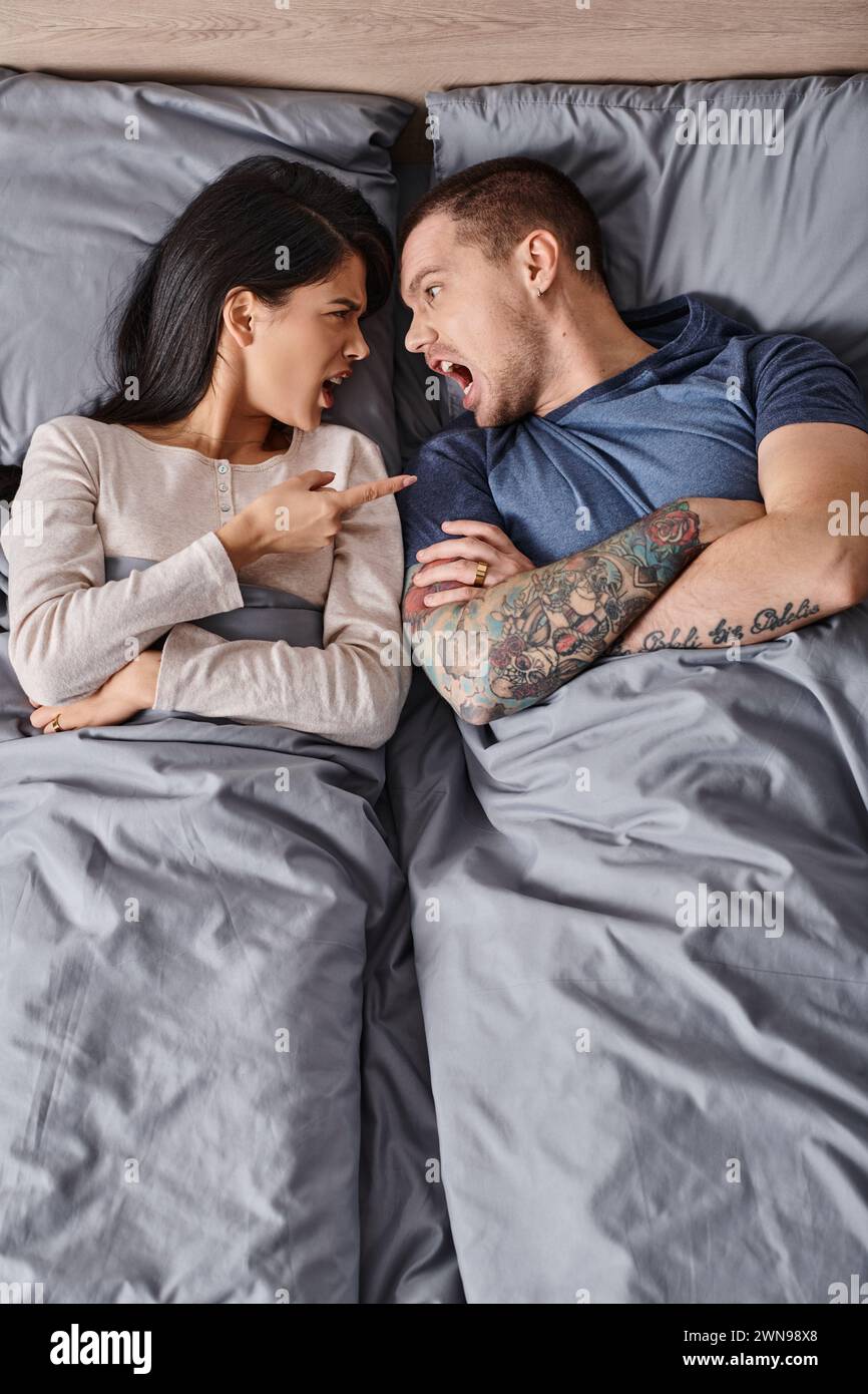top view of angry multicultural couple lying down on bed screaming and ...