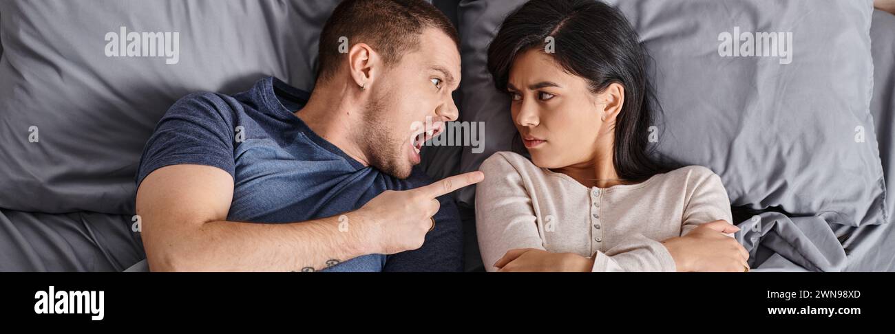 angry multicultural couple lying down on bed shouting and quarreling at ...