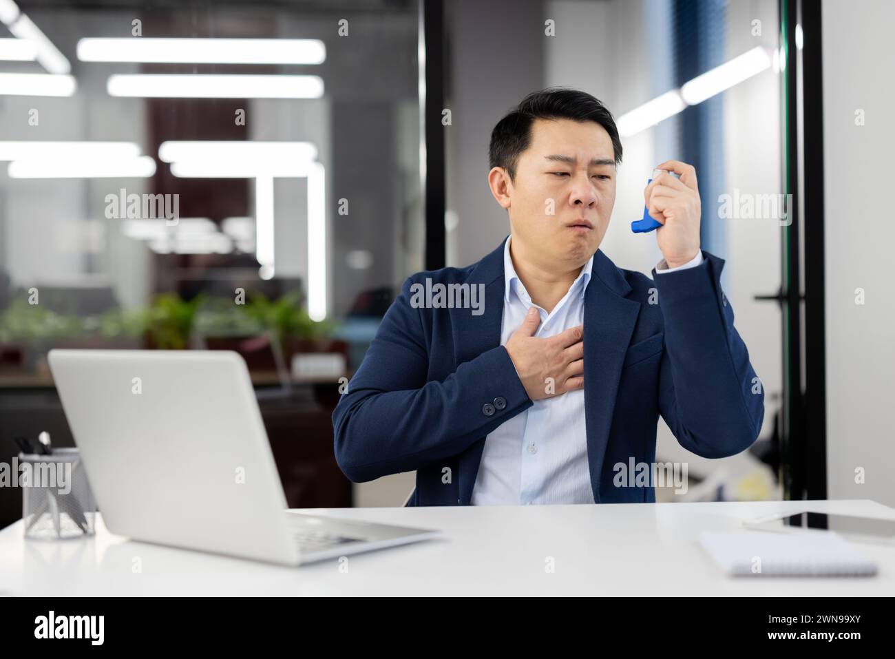 Sick japanese ceo grabbing chest with hand and holding medicine inhaler while sitting by table ...