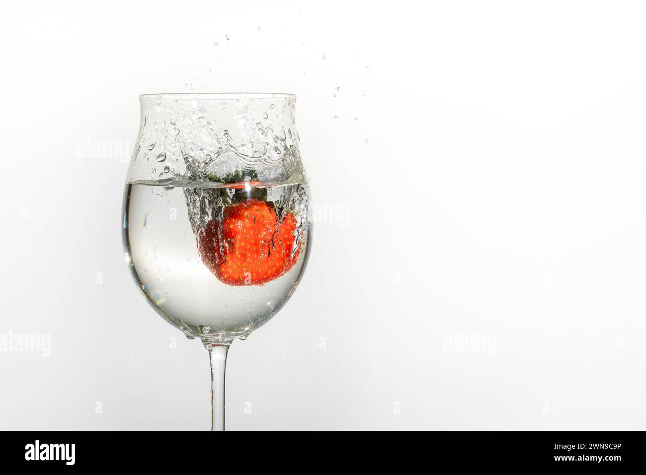 A strawberry falls into a wine glass, splashing water against a white ...