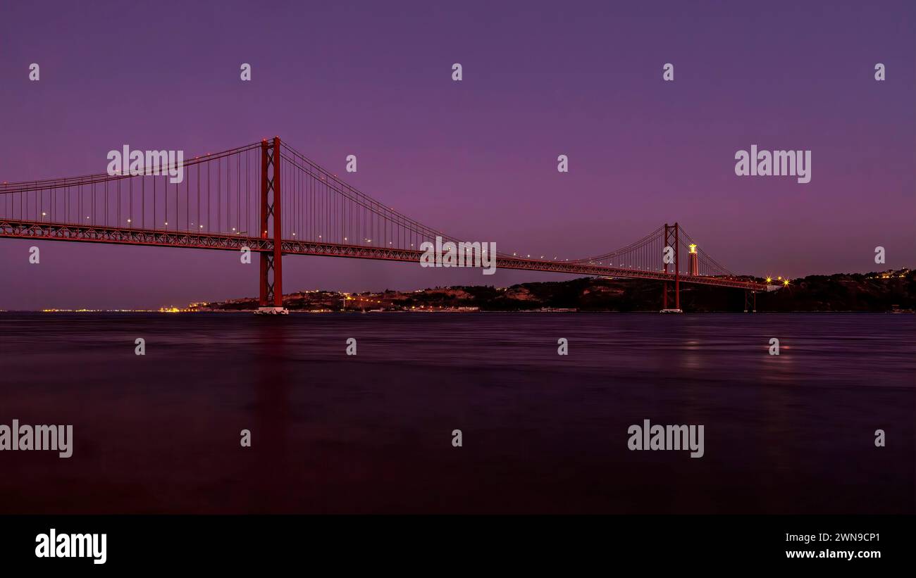 Twilight descends on a calm river with a suspension bridge standing ...