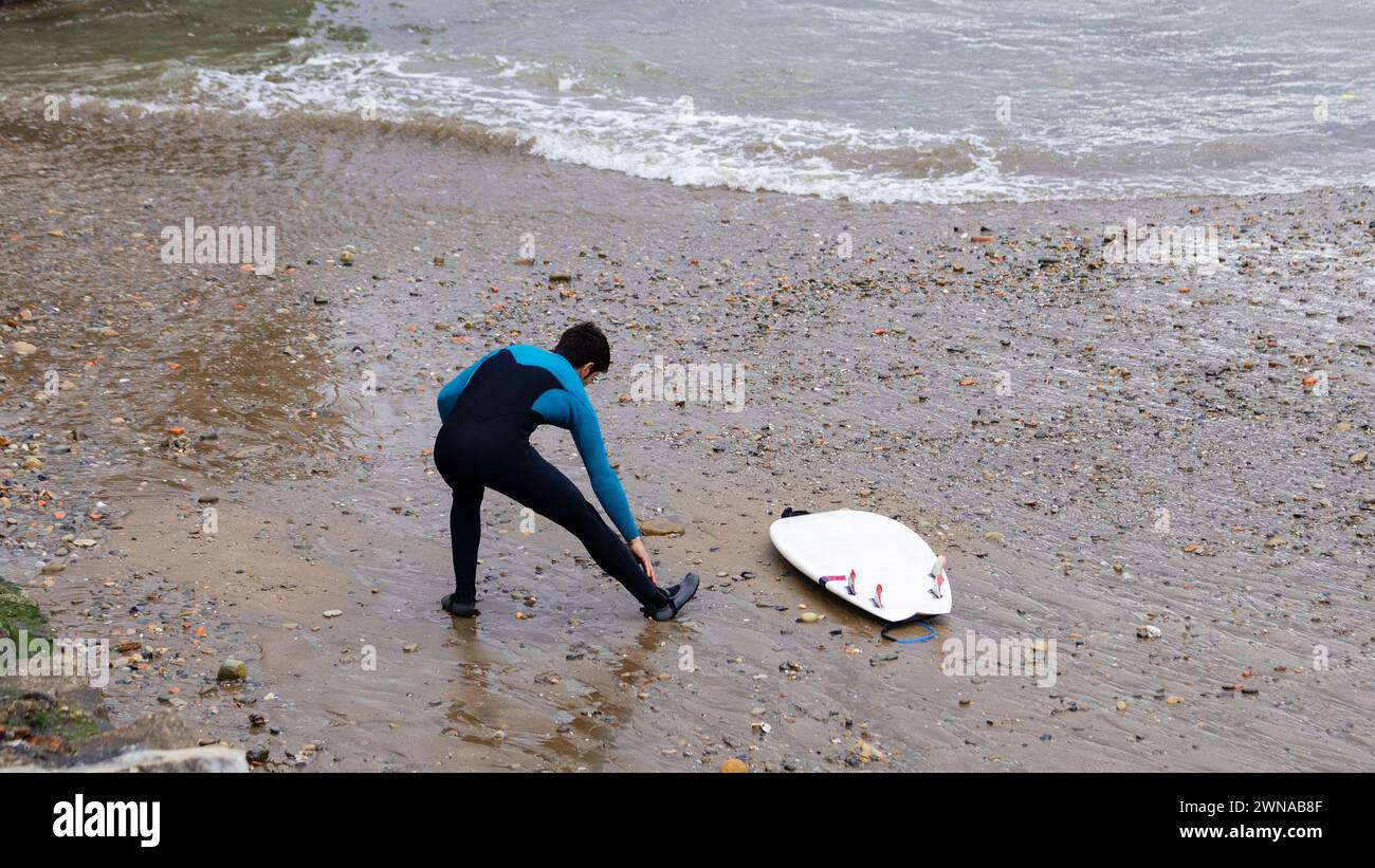 A surfer in a blue wetsuit secures his suit on a pebbly shore before a ...