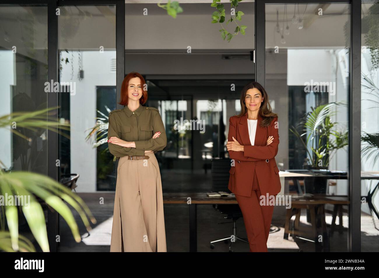 Two smiling confident ladies standing arms crossed in office, portrait ...