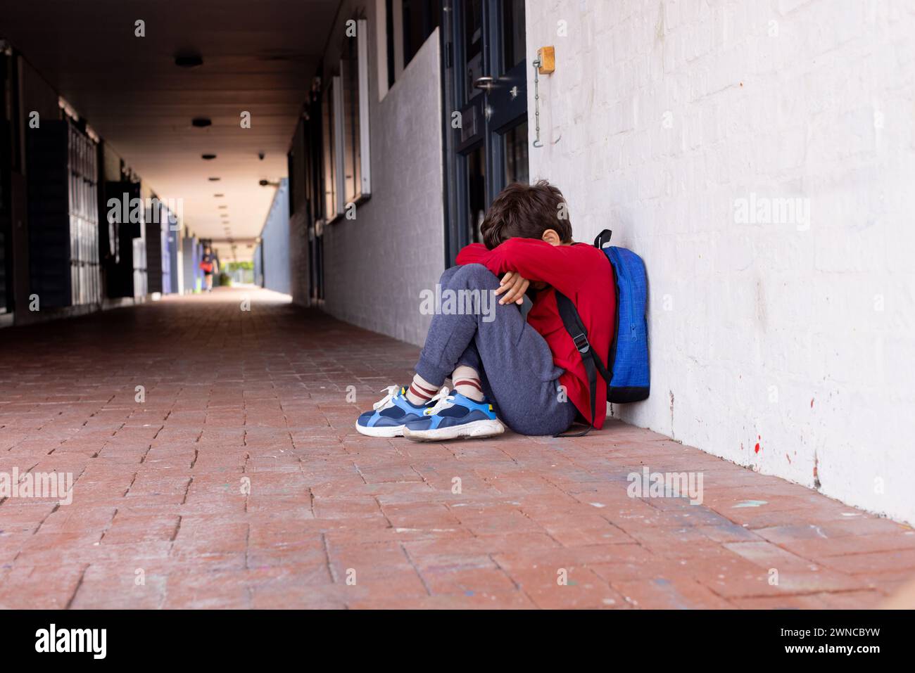 A biracial boy sits alone in a school corridor, with a red backpack ...