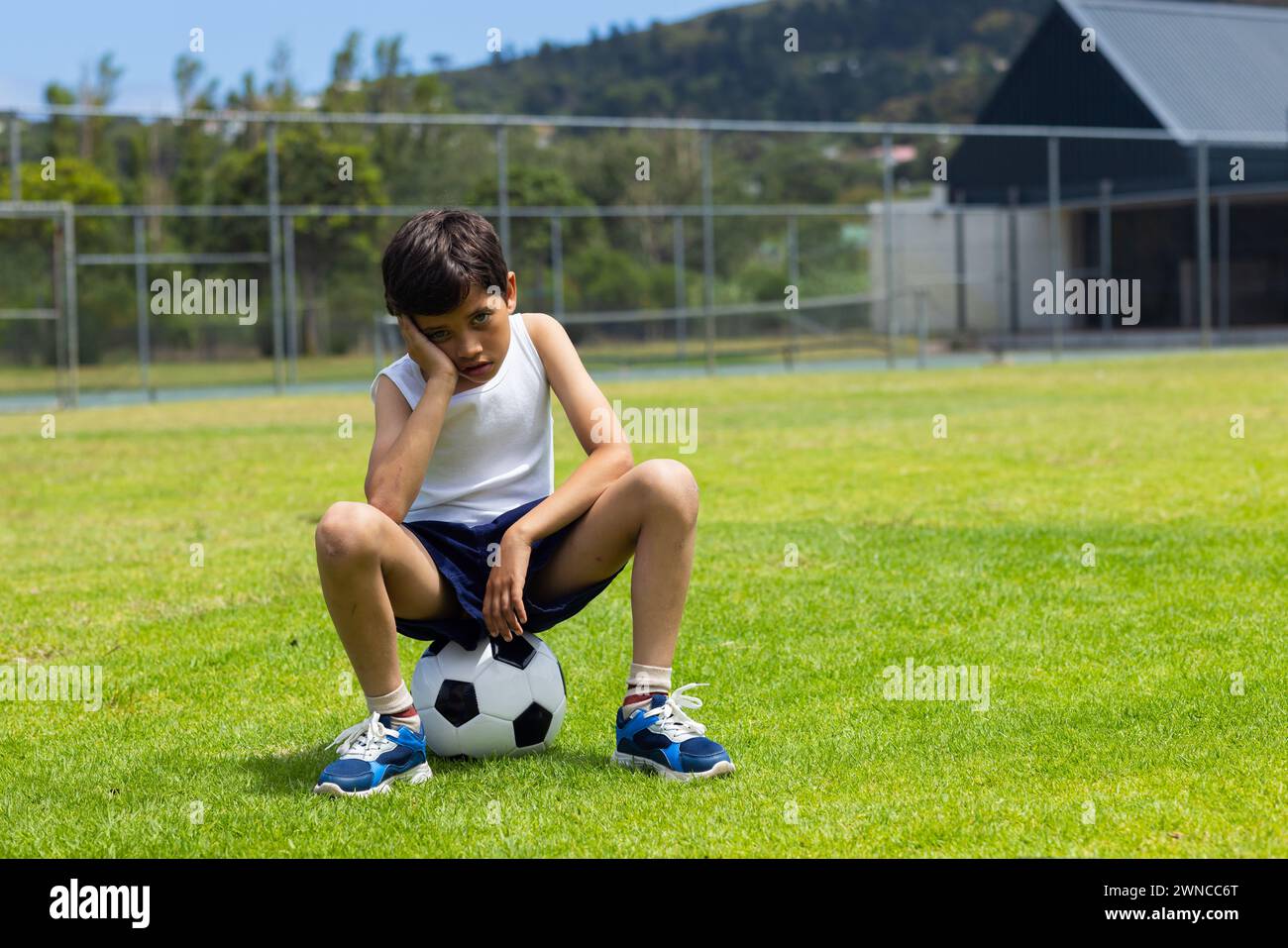 Biracial boy sits on a soccer ball, looking sad on a sunny field at ...