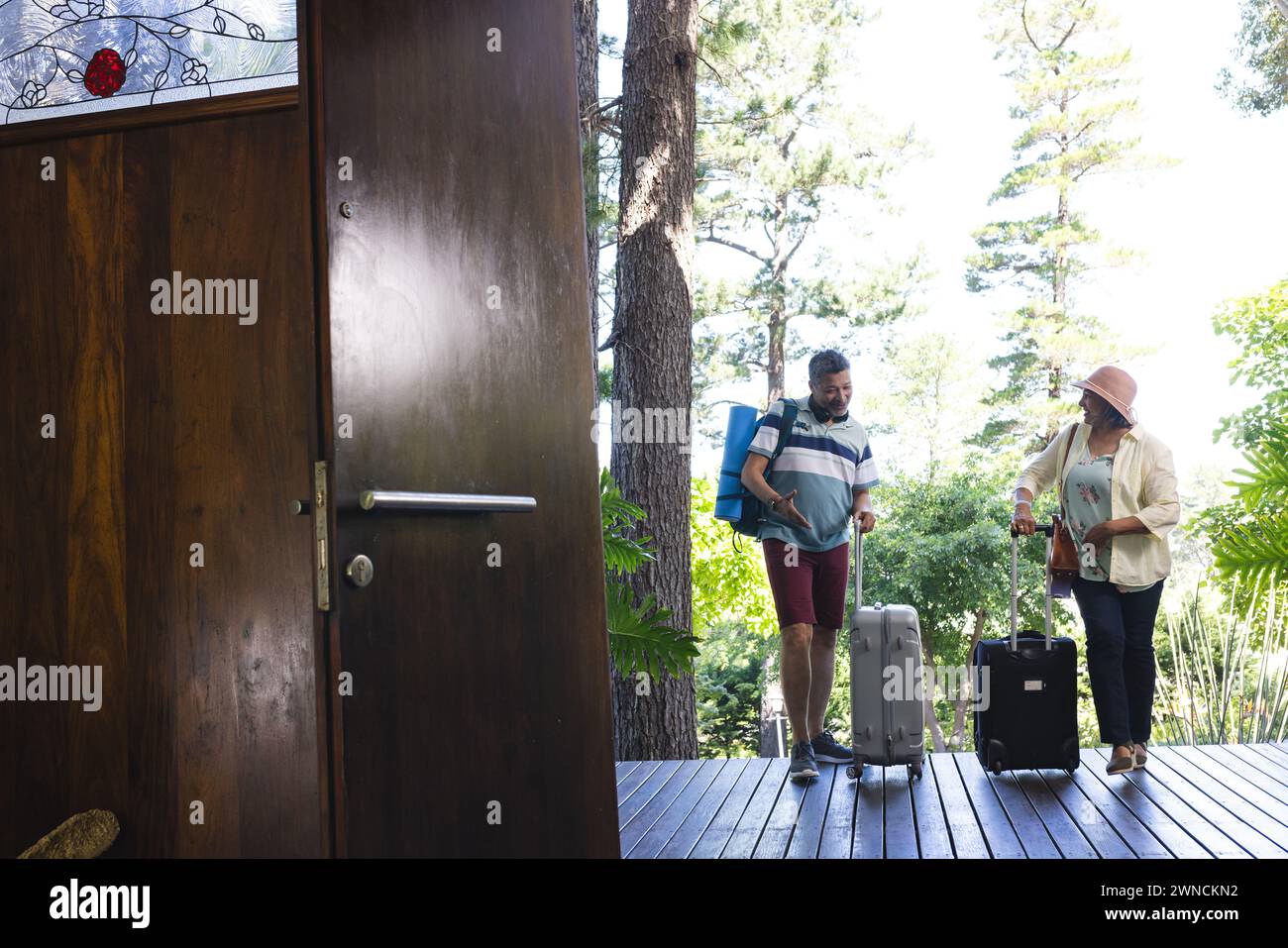 Senior biracial couple with luggage at a cabin, surrounded by trees ...