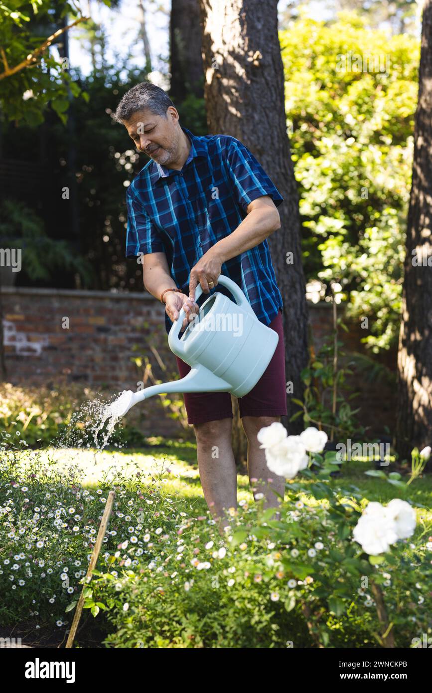 Senior biracial man waters plants in a sunny garden, wearing a blue ...