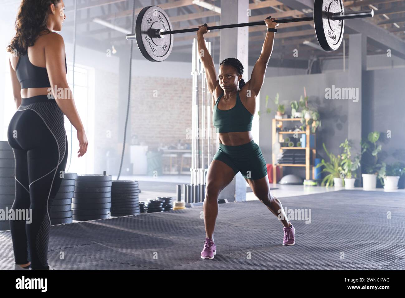 Young strong biracial woman lifts weights at a gym, observed by a ...