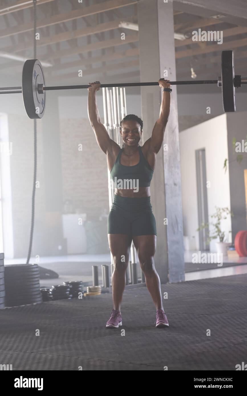 Strong biracial woman lifts a barbell overhead in a gym setting ...