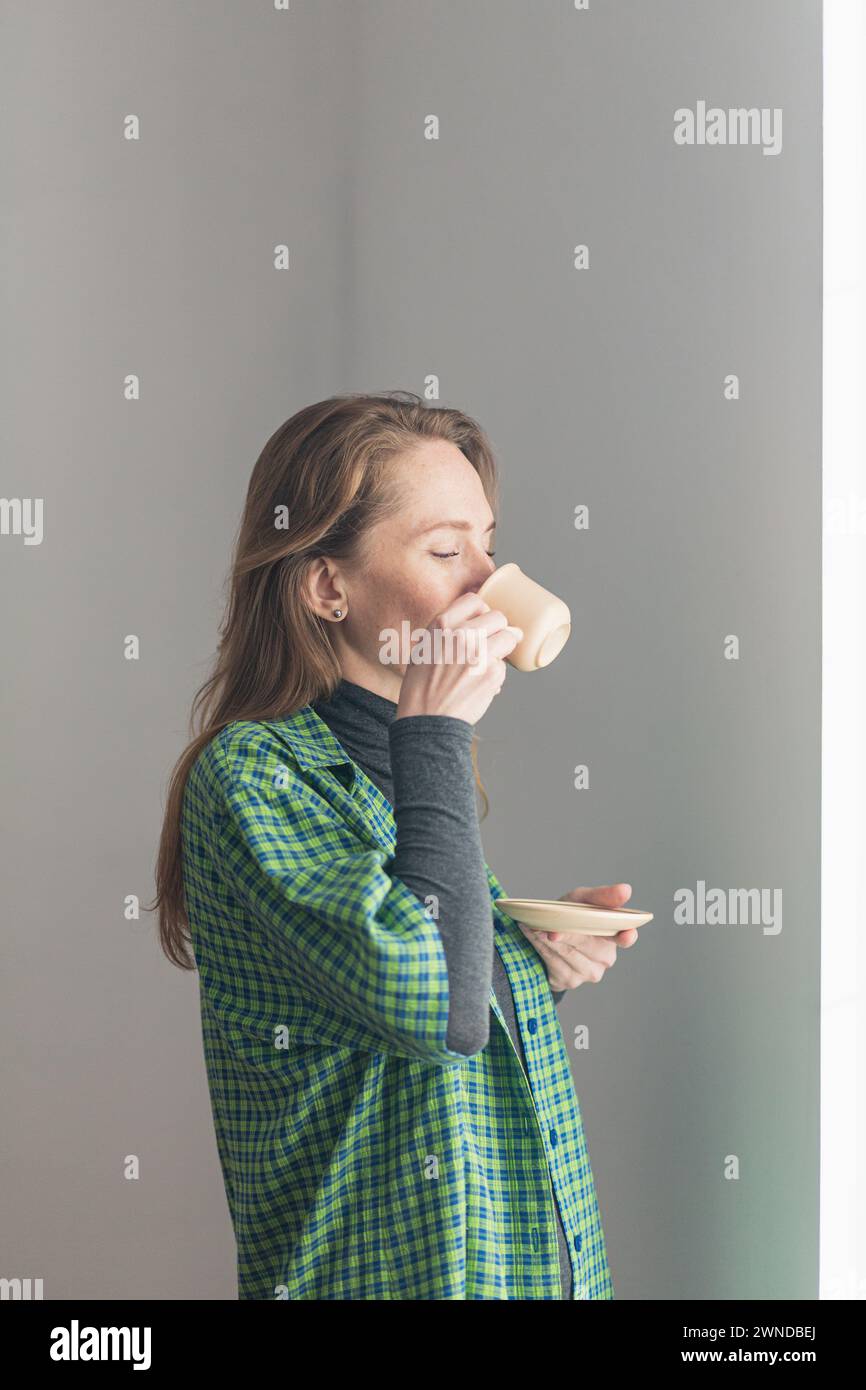 A young adult woman stands near the window and drinks from a cup ...