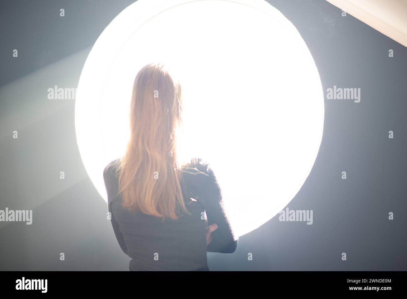 A woman stands facing the window, bathed in the rays of the sun. The ...