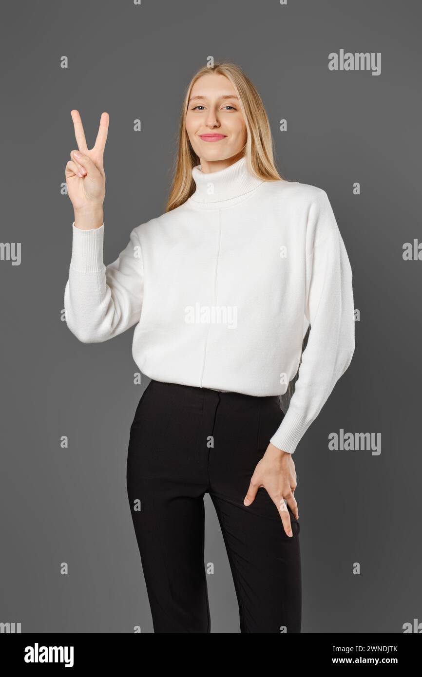 Young woman in trousers and sweater shows victory sign lifting two ...