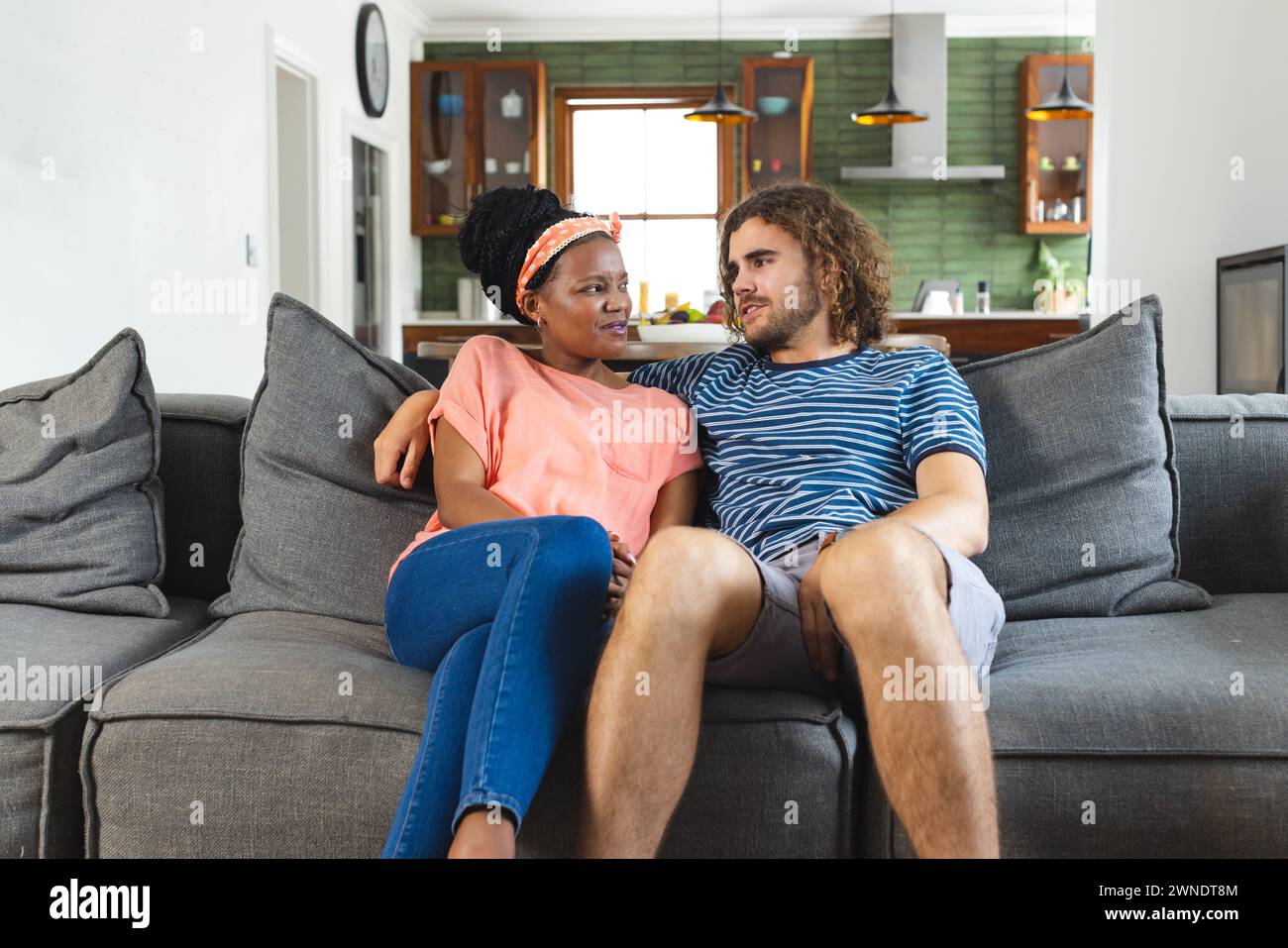 Diverse couple sitting closely on a grey sofa, sharing a moment of ...