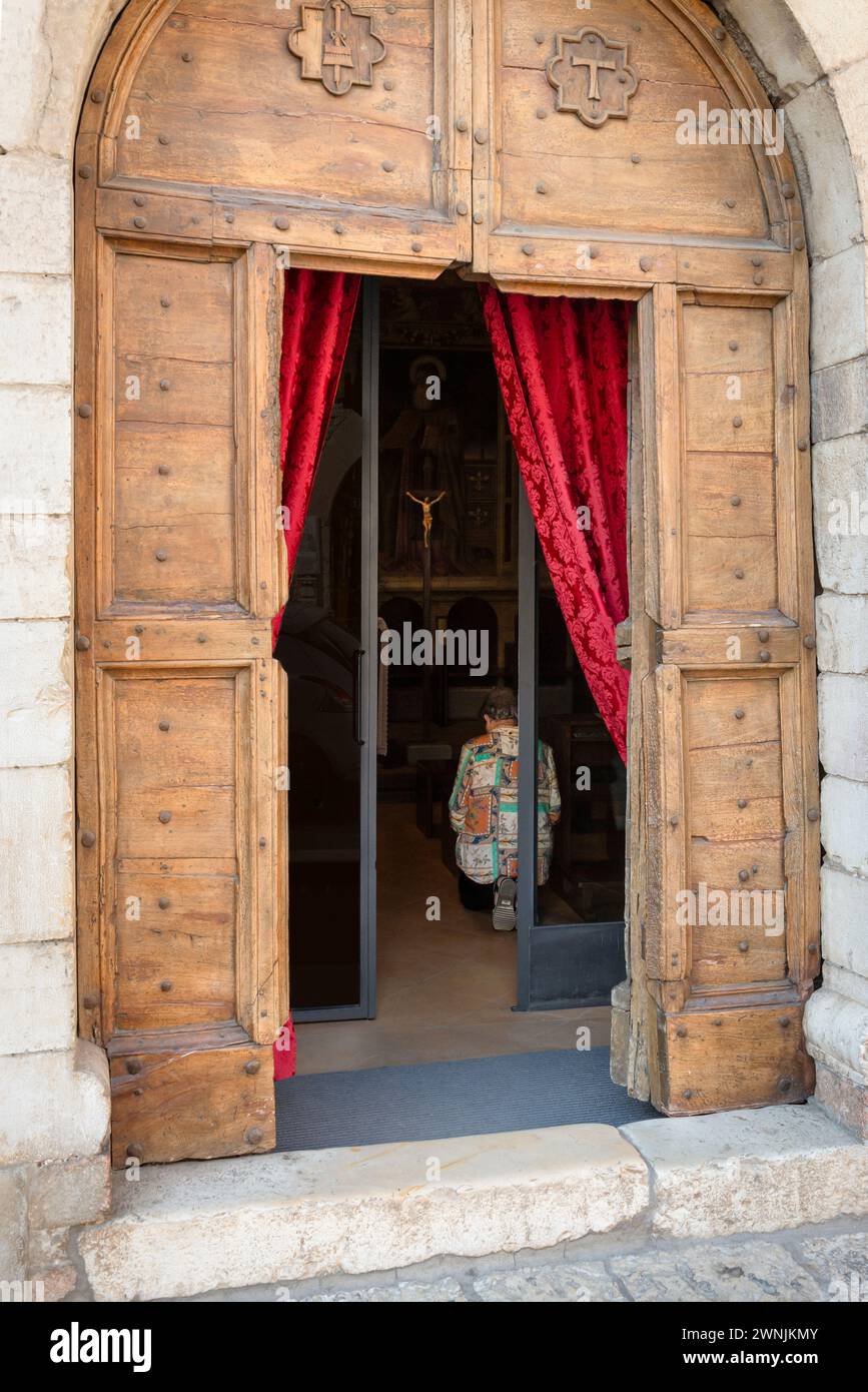 An elderly woman kneels behind a wooden door in a chapel in front of an ...