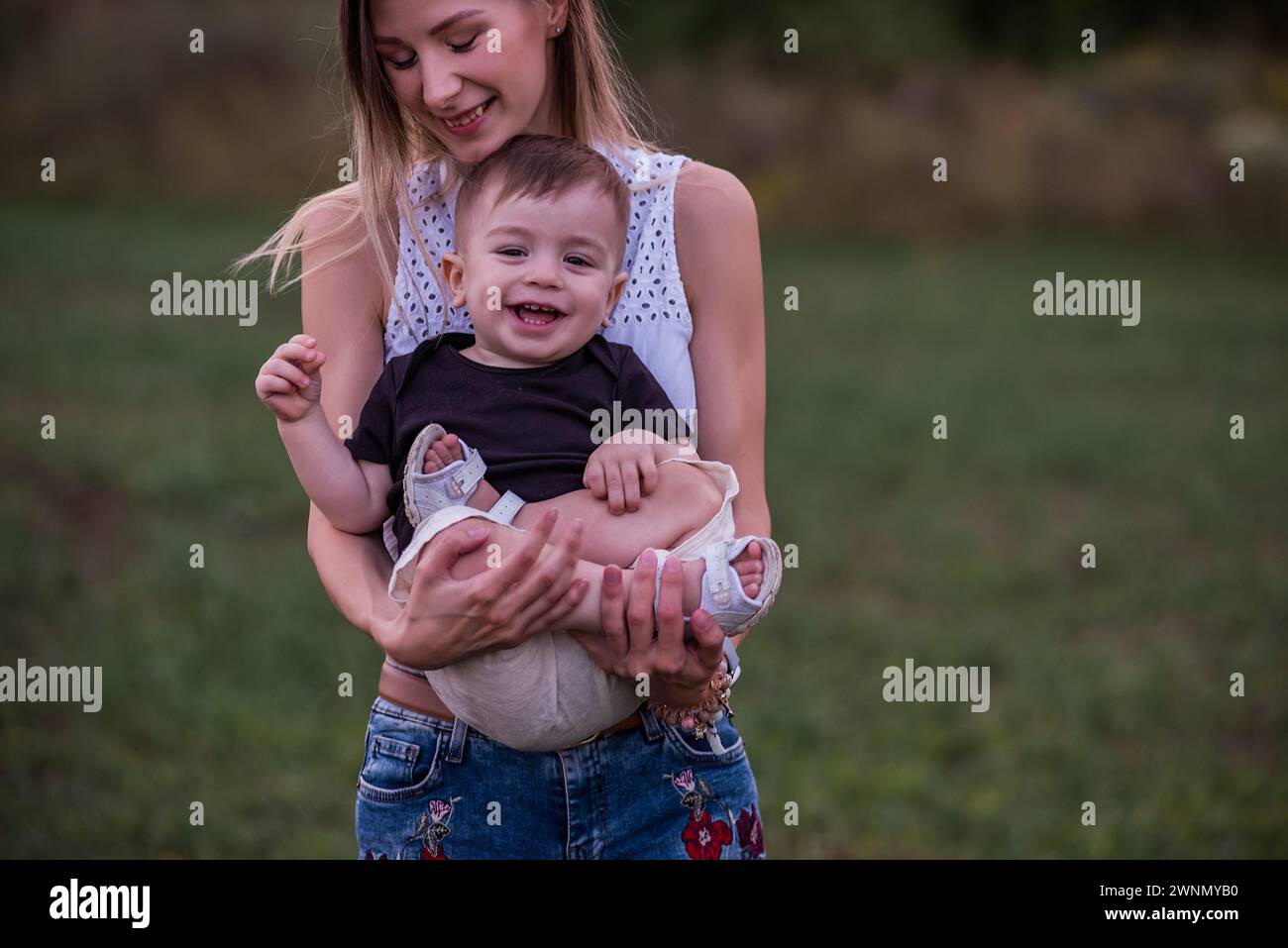 Young woman smiles, holds toddler son in her arms, standing in lush ...