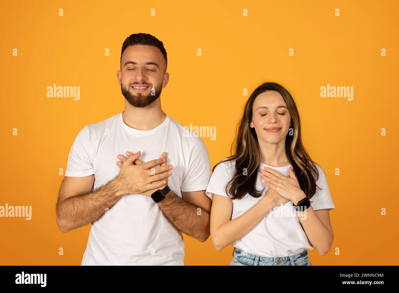 Grateful young couple with eyes closed and hands on hearts, expressing ...