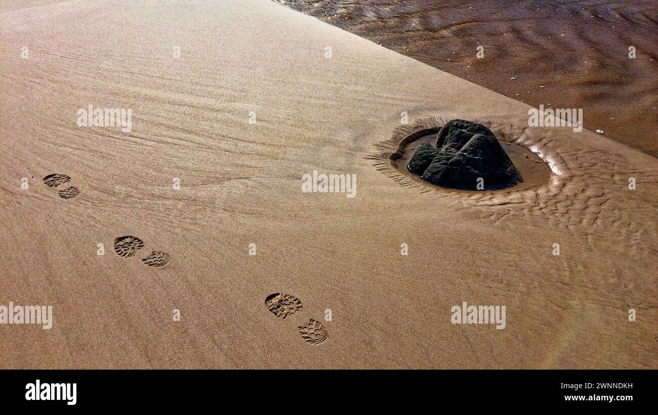 Footprints on wet sand head towards a dark, wet rock, with patterns ...