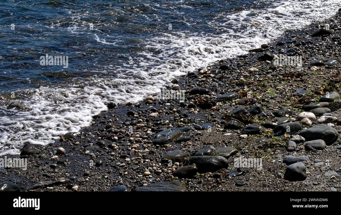 The image shows a rocky shoreline with waves crashing. The pebbles and ...