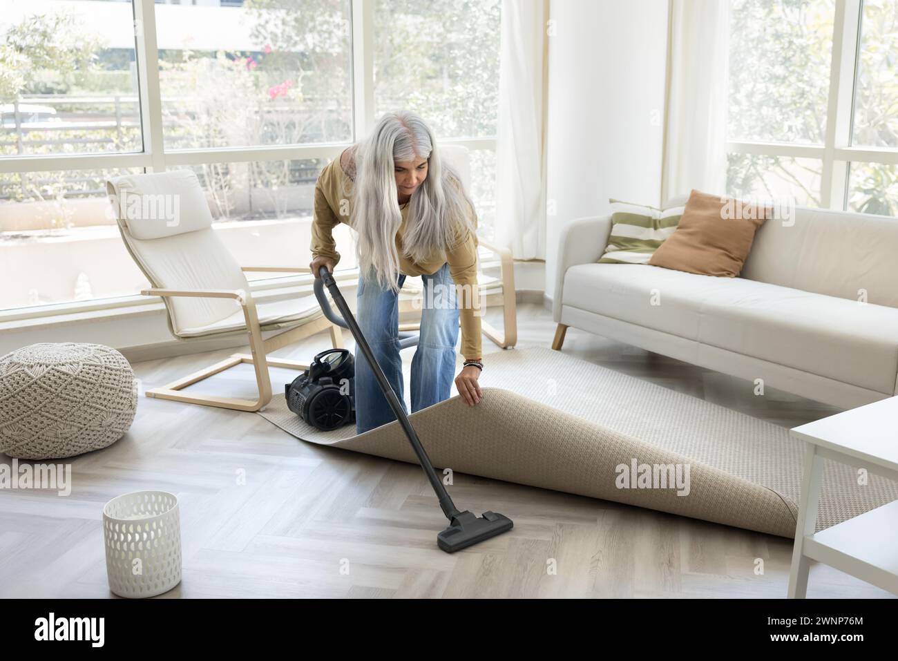 Serious grey haired mature housemaid woman hoovering floor under rug ...