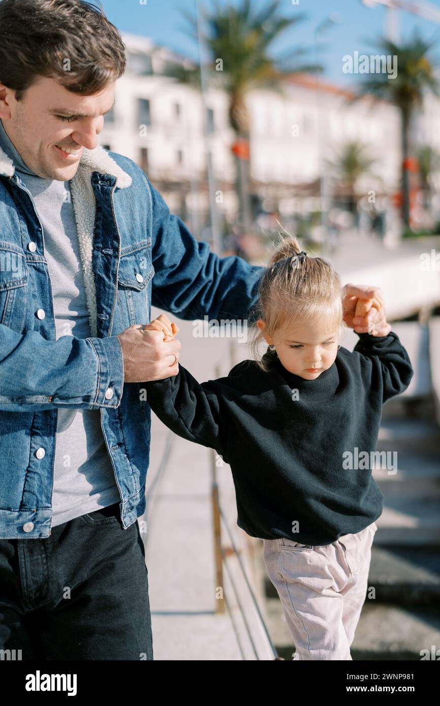 Dad holds the hands of a little girl walking along a rope fence on the ...