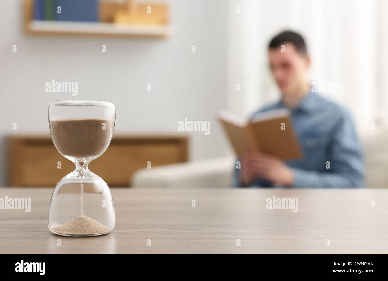Hourglass with flowing sand on desk. Man reading book in room ...